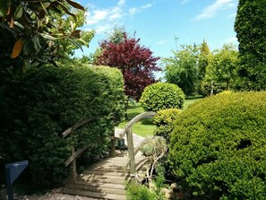 Uma passarela de madeira serpenteia por um jardim bem cuidado, com arbustos diversos e uma grande árvore de folhas vermelhas.