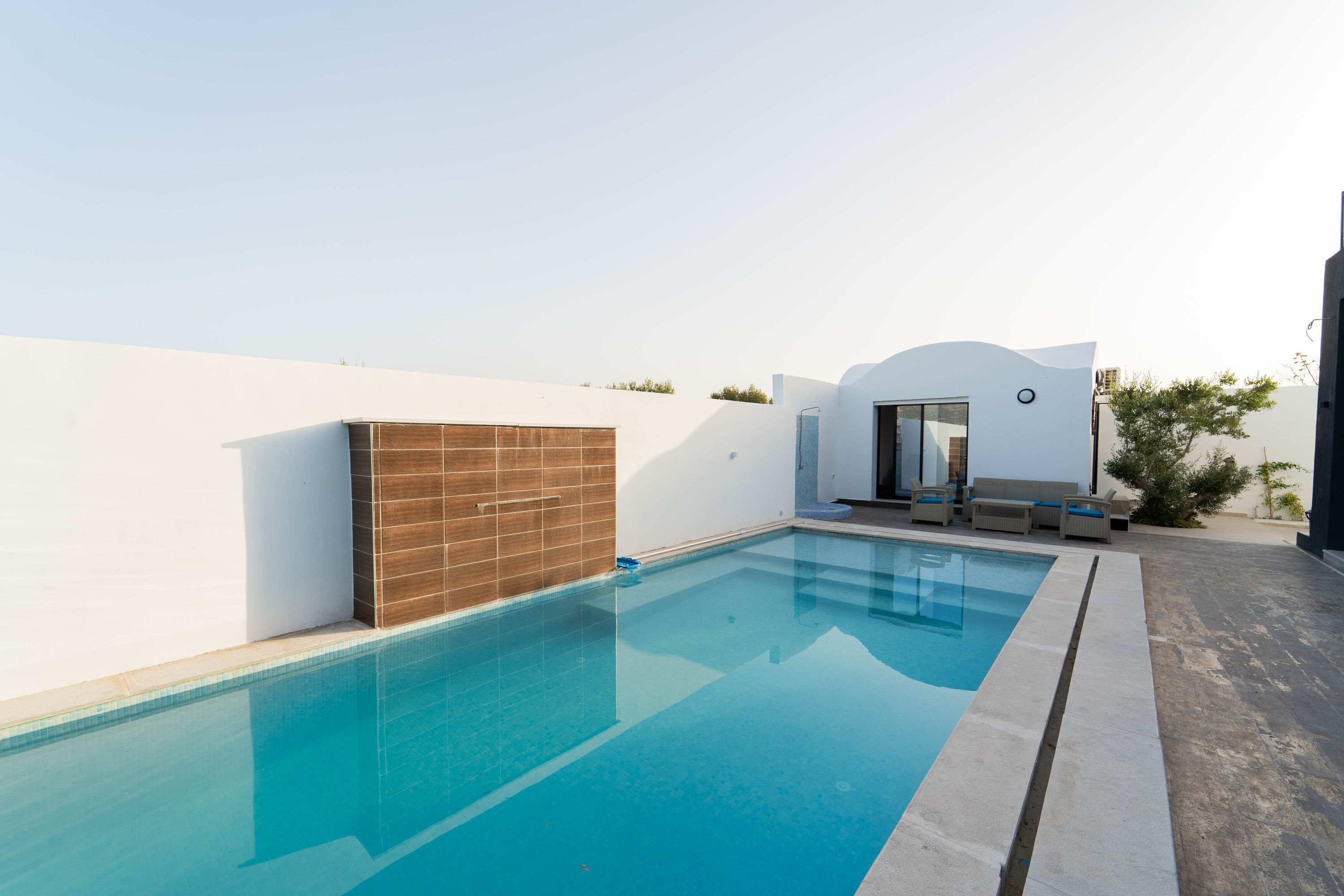 Uma piscina com um deck revestido de azulejos, uma parede branca e uma pequena área de estar ao ar livre com cadeiras e uma mesa.