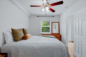 Quarto com cama, almofadas, mesa de cabeceira, cômoda, janela com persianas e ventilador de teto.