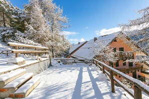 Área externa coberta de neve, com bancos de madeira, parede de pedra e um edifício com telhado inclinado.