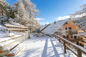 Área externa coberta de neve, com bancos de madeira, parede de pedra e um edifício com telhado inclinado.