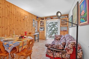 Interior de madeira com mesa de jantar posta, um sofá e vista para árvores cobertas de neve através de uma porta de vidro.