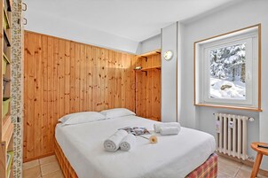 Quarto com parede de madeira, cama com roupa de cama branca, janela com vista nevada e radiador.