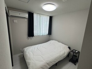 Quarto pequeno com uma cama de solteiro, edredom branco, janela com cortinas leves, ar condicionado na parede e uma mesa com um cesto de lixo.