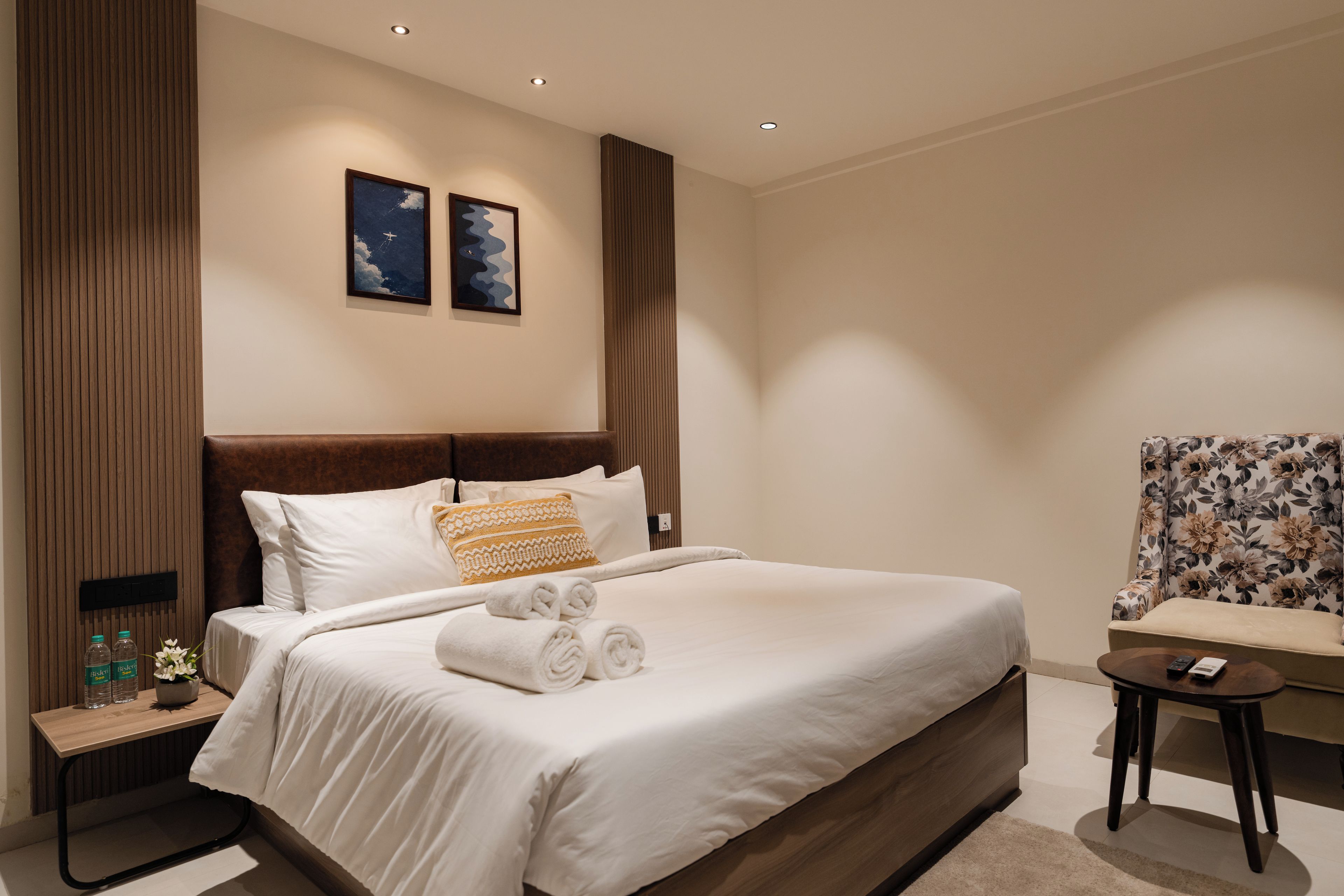 Quarto de hotel com uma cama grande, uma mesa de cabeceira com abajur, uma cadeira e duas fotos emolduradas na parede.
