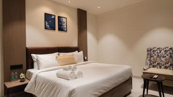 Quarto de hotel com uma cama grande, uma mesa de cabeceira com abajur, uma cadeira e duas fotos emolduradas na parede.