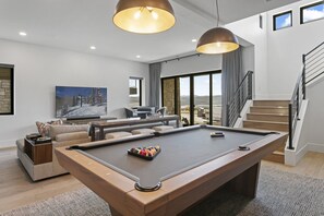 Uma sala de estar moderna com mesa de bilhar, um conjunto de sofás e uma grande janela com vista para o exterior.