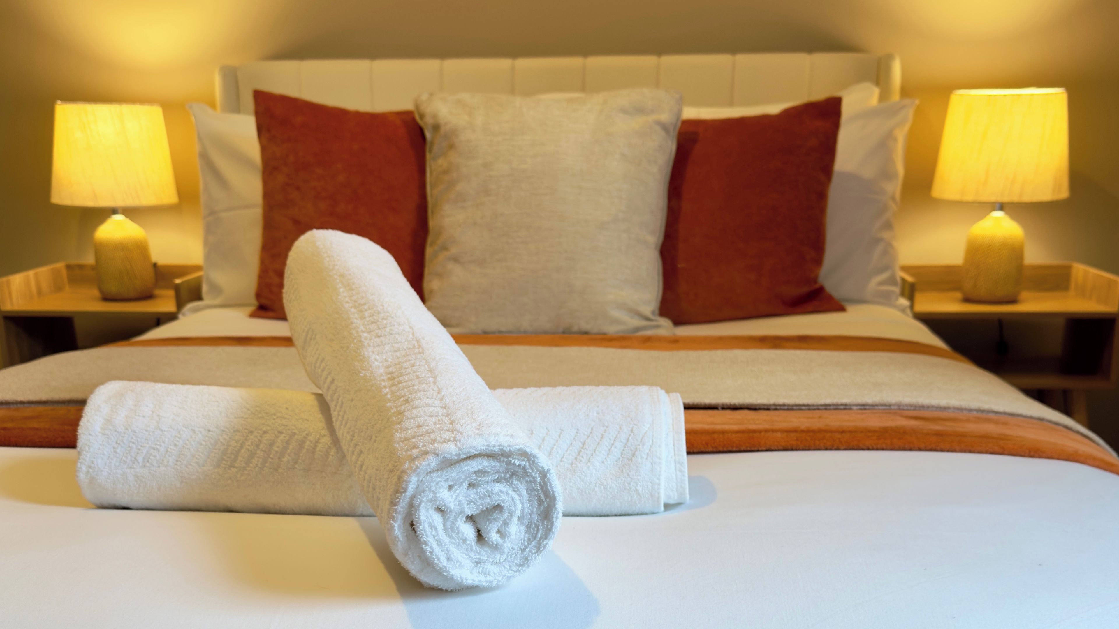 Cama bem arrumada com toalha enrolada, duas luminárias de cabeceira e cabeceira com dois travesseiros.