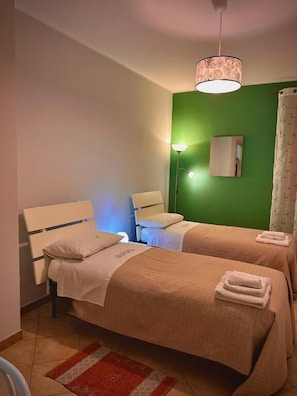 Quarto de hotel com duas camas, uma parede de destaque verde, uma luminária pendente e uma mesinha de cabeceira.