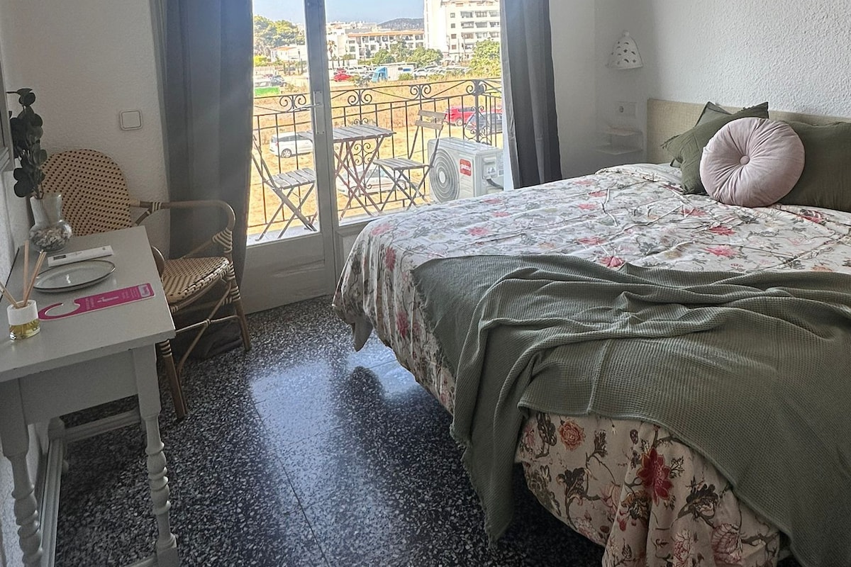 Quarto de hotel com cama, uma mesinha, uma cadeira, janela com cortinas e aparelho de ar condicionado.