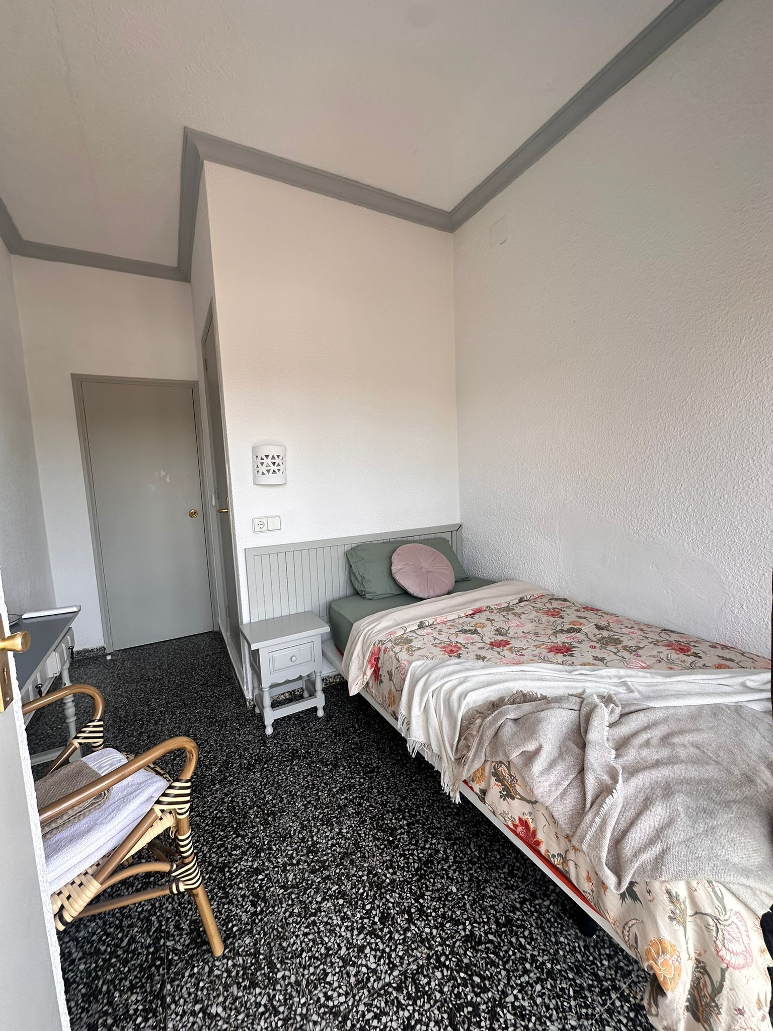 Quarto pequeno e compacto com uma cama de solteiro, mesa de cabeceira, uma cadeira e porta que dá para outro cômodo.