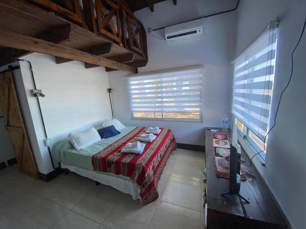 Quarto com cama, janela com persianas, escrivaninha com computador e ventilador de teto.