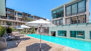 Piscina com deck de madeira, guarda-sóis e um edifício com varandas ao fundo.