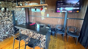 Uma área de bar com parede de pedra, teto de madeira e uma televisão de tela plana exibindo um canal de notícias.