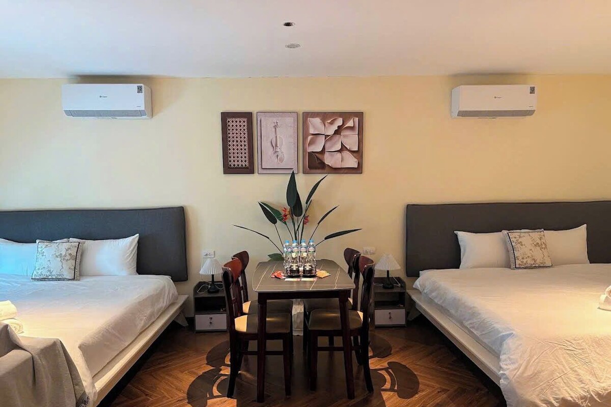 Quarto de hotel com duas camas, uma mesa de jantar e cadeiras, e obras de arte nas paredes.