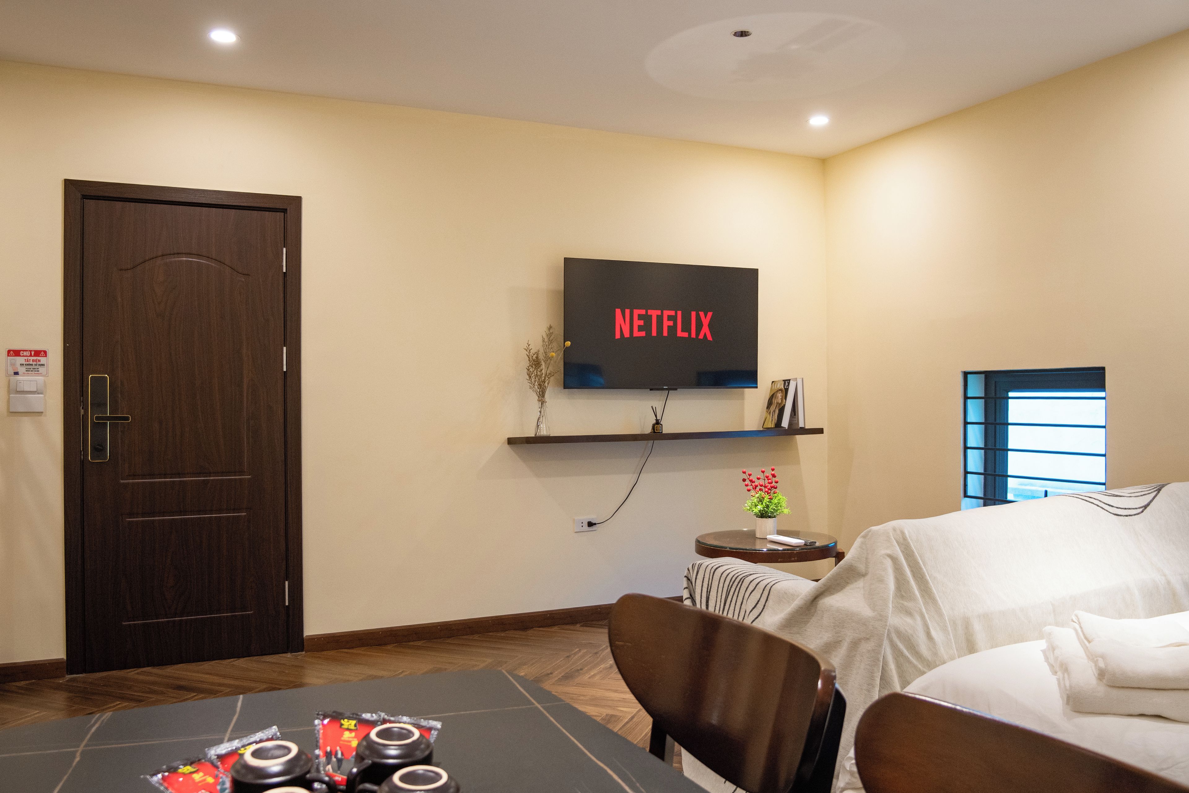 Quarto de hotel com cama, televisão na parede, uma mesinha com lanches e uma porta.