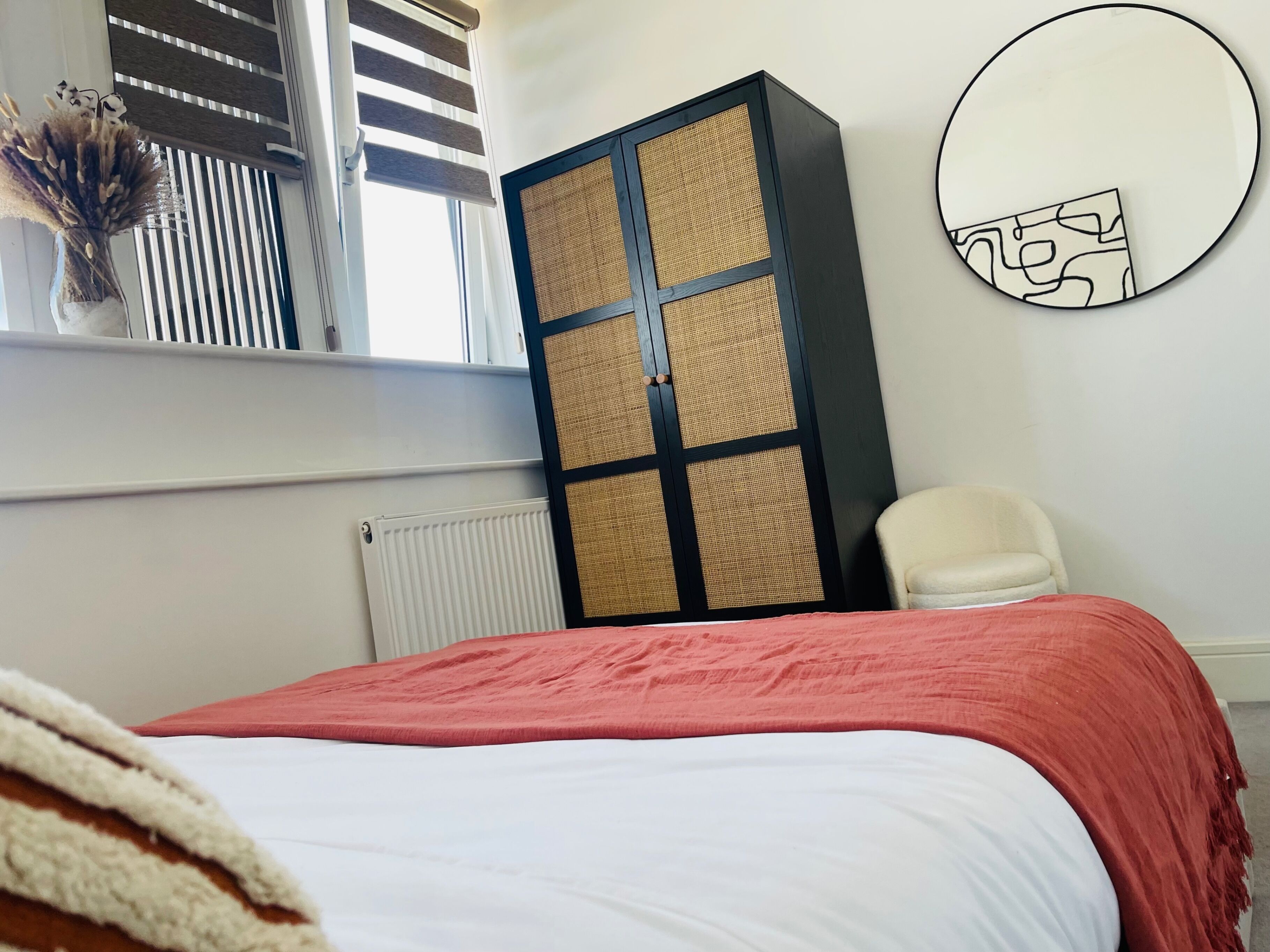 Quarto com cama, radiador, janela com persianas, cadeira e espelho fixado na parede.