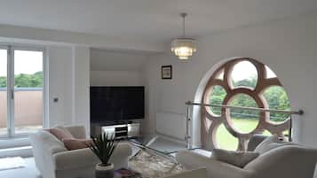 Uma sala de estar moderna com televisão de tela plana, assentos confortáveis, uma mesa de centro de vidro e uma ampla janela em arco com vista para vegetação.