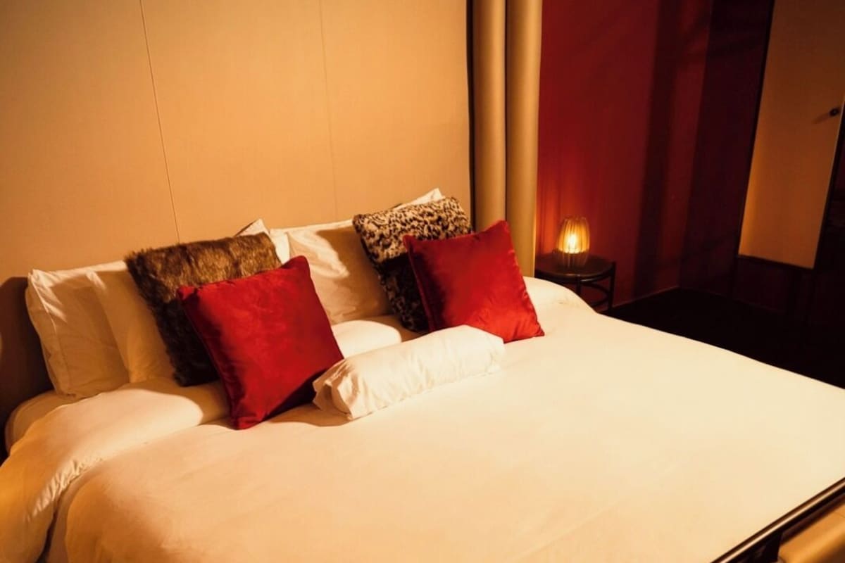 Cama com roupa de cama branca e várias almofadas, incluindo algumas vermelhas e estampadas, em um quarto com iluminação fraca, mesa de cabeceira e abajur.