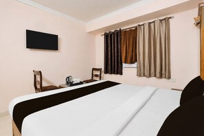 Quarto de hotel com cama, televisão na parede, janela com cortinas e pequena mesa com chaleira e chávenas.