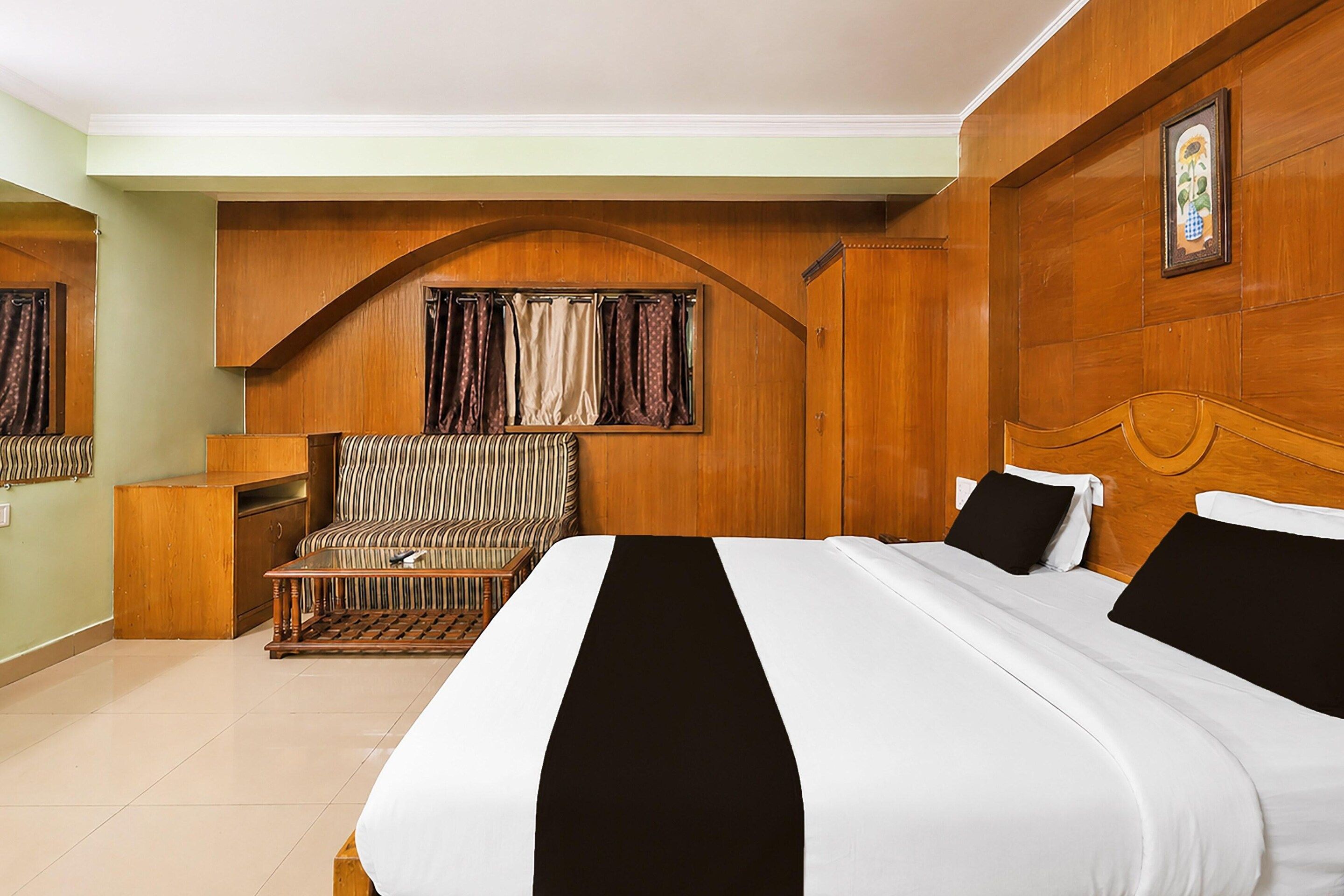 Quarto de hotel com uma cama grande, um banquinho de madeira, uma mesa pequena e um espelho.
