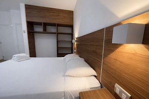 Quarto de hotel moderno com cama bem arrumada, cabeceira de madeira, roupeiro embutido e mesa de cabeceira com abajur.