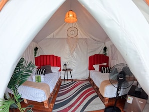 Acomodação em tenda com duas camas, uma mesinha, uma luminária pendente e um ventilador.