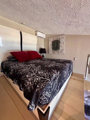Quarto com cama, travesseiro vermelho, luminária preta e ar-condicionado fixado na parede.