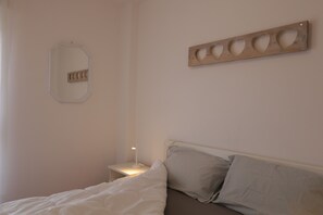 Quarto com cama, mesa de cabeceira com abajur, espelho fixado na parede e uma obra de arte em madeira.