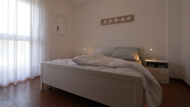 Quarto com cama, mesa de cabeceira, espelho fixado na parede e janela com cortinas translúcidas.