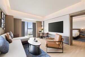Quarto de hotel moderno com sofá, poltrona, TV de tela plana e vista para a cidade.