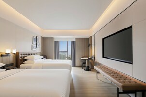 Quarto de hotel com duas camas, uma televisão grande, um banco e vista para a cidade.