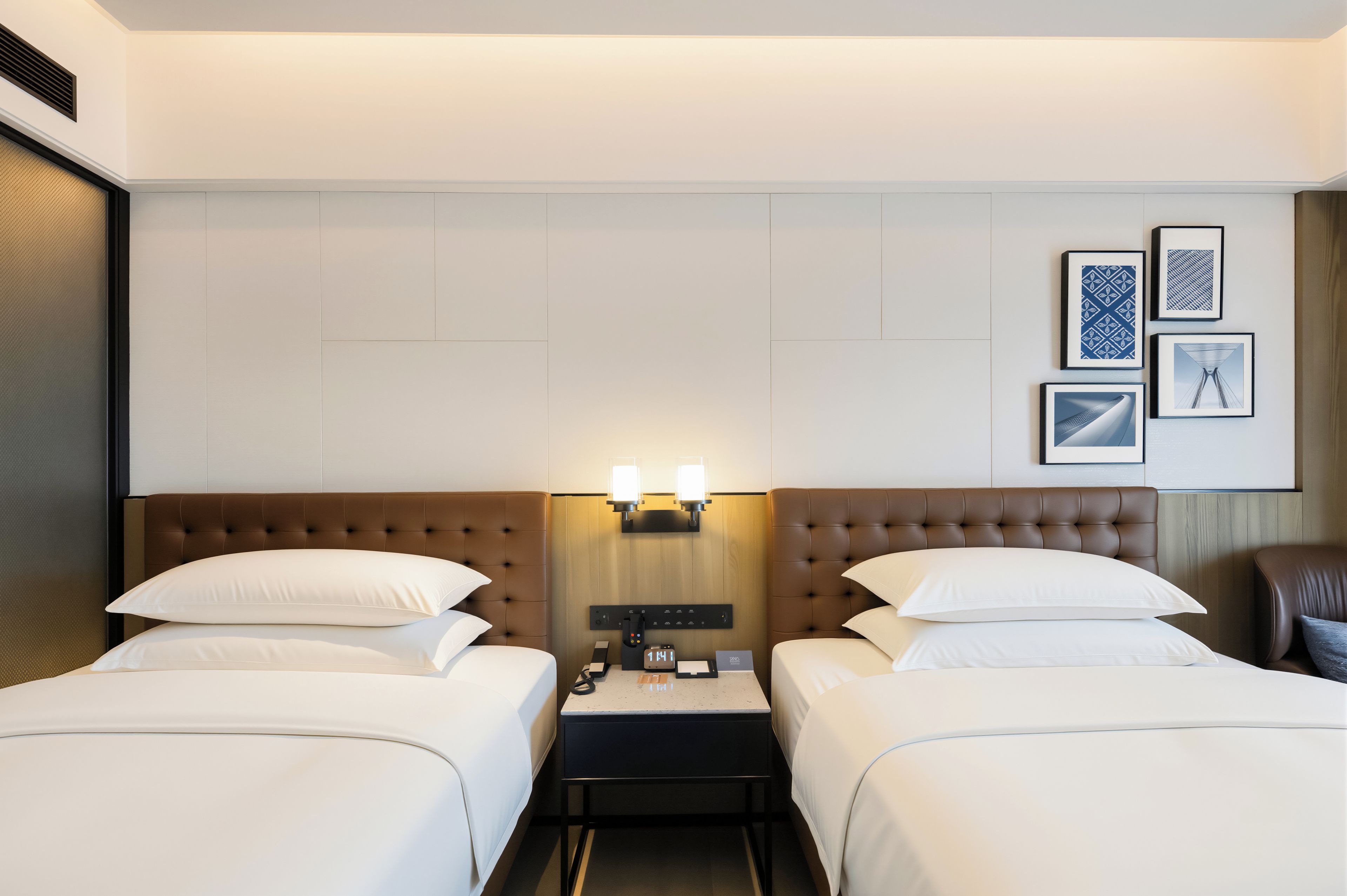 Quarto de hotel com duas camas, mesa de cabeceira, luminária na parede e quadros decorativos.