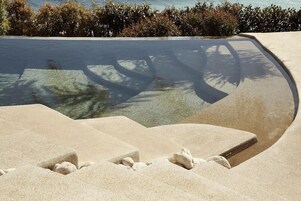 Uma piscina com água cristalina, rodeada por areia e vegetação.