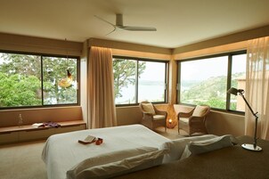 Quarto de hotel com cama, duas cadeiras de vime, uma mesa e janelas amplas com vista para o exterior.