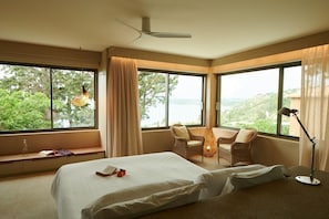 Quarto de hotel com cama, duas cadeiras de vime, uma mesa e janelas amplas com vista para o exterior.