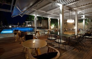 Área de piscina ao ar livre com deck de madeira, mesas e cadeiras, cercada por um alpendre coberto com vista para a piscina.