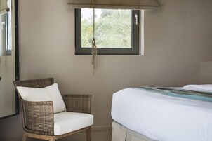 Quarto com cama, uma cadeira de vime e janela com vista para o verde.