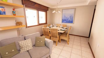 Uma área de refeições com uma mesa posta para quatro pessoas, rodeada por cadeiras de vime, um sofá com almofadas e uma janela com persianas.