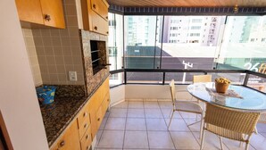 Uma cozinha com armários de madeira e forno embutido, uma sala de jantar com mesa de vidro e cadeiras de vime, e uma varanda com vista para edifícios.