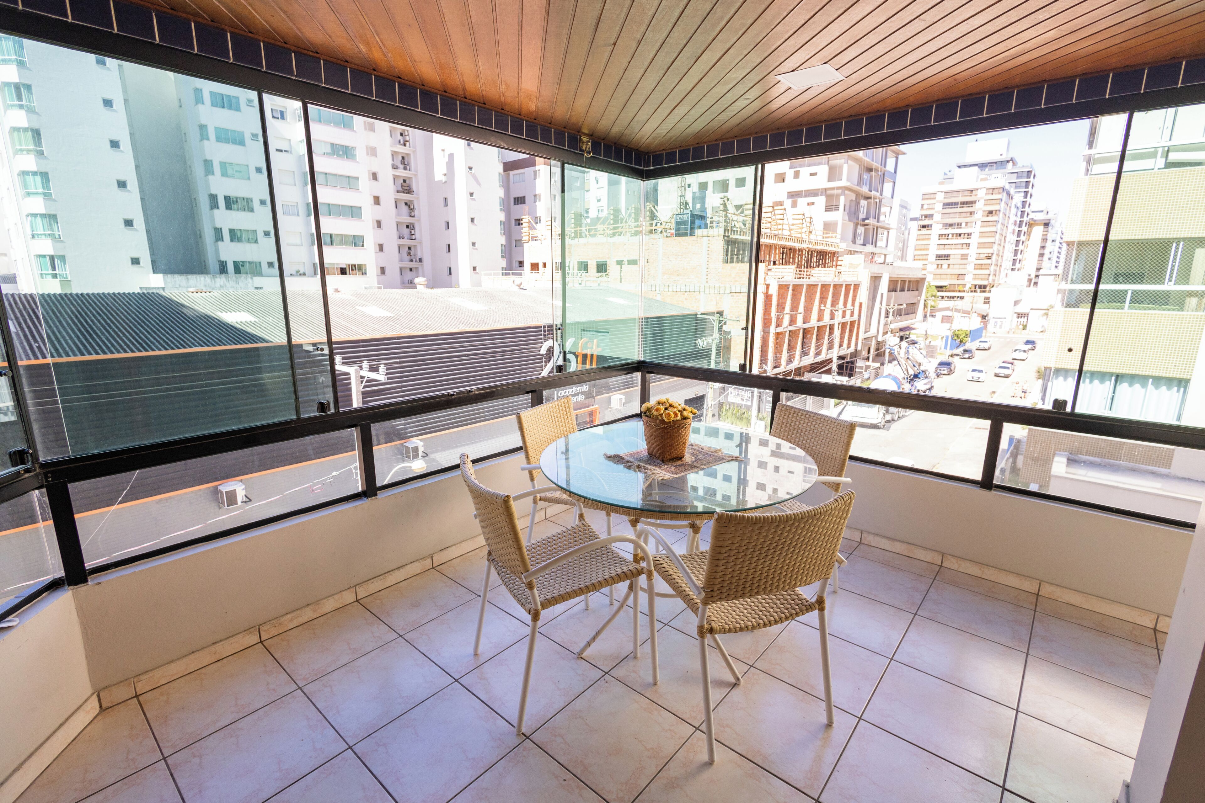 Uma varanda com uma mesa de vidro redonda e cadeiras de vime, com vista para uma paisagem urbana com edifícios e um canteiro de obras.