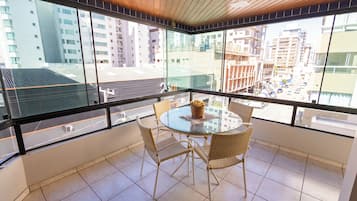 Uma varanda com uma mesa de vidro redonda e cadeiras de vime, com vista para uma paisagem urbana com edifícios e um canteiro de obras.