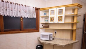 Uma cozinha compacta com micro-ondas, lixeira e janela com cortina de renda.