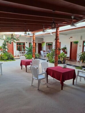 Área de estar ao ar livre com mesas e cadeiras, cercada por plantas e colunas.
