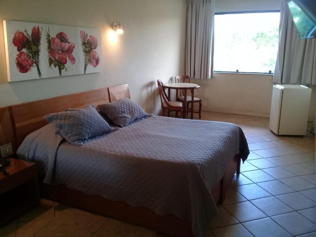 Um quarto com cama, mesa de cabeceira, uma mesa com cadeiras e janela com cortinas.