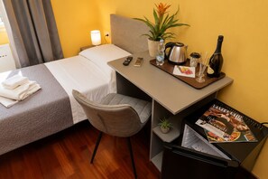 Quarto de hotel com cama, secretária com cadeira, pequena mesa com planta, chaleira, garrafa e revista.