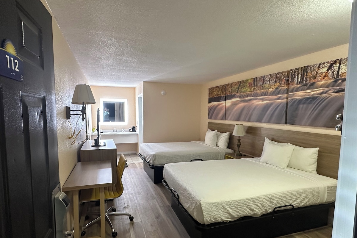 Quarto de hotel com duas camas, uma escrivaninha, uma cadeira, uma janela e uma obra de arte na parede.
