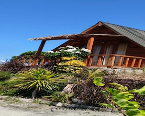 Uma cabana de madeira com telhado de palha, rodeada por plantas tropicais e um céu azul límpido.