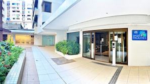 Um edifício moderno com entrada de vidro, uma placa com 'Maceió Facilities' e um caminho de azulejos com plantas.