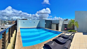 Uma piscina no terraço com água azul cristalina, rodeada por um deck de azulejos e espreguiçadeiras.